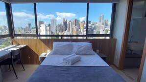 Quarto com vista para a cidade, uma escrivaninha e uma cama com roupa de cama branca.