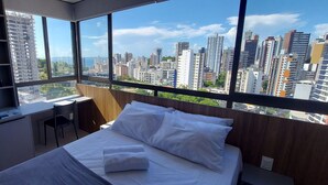 Quarto de hotel moderno com uma cama grande, uma escrivaninha e vista para o horizonte urbano pelas amplas janelas.