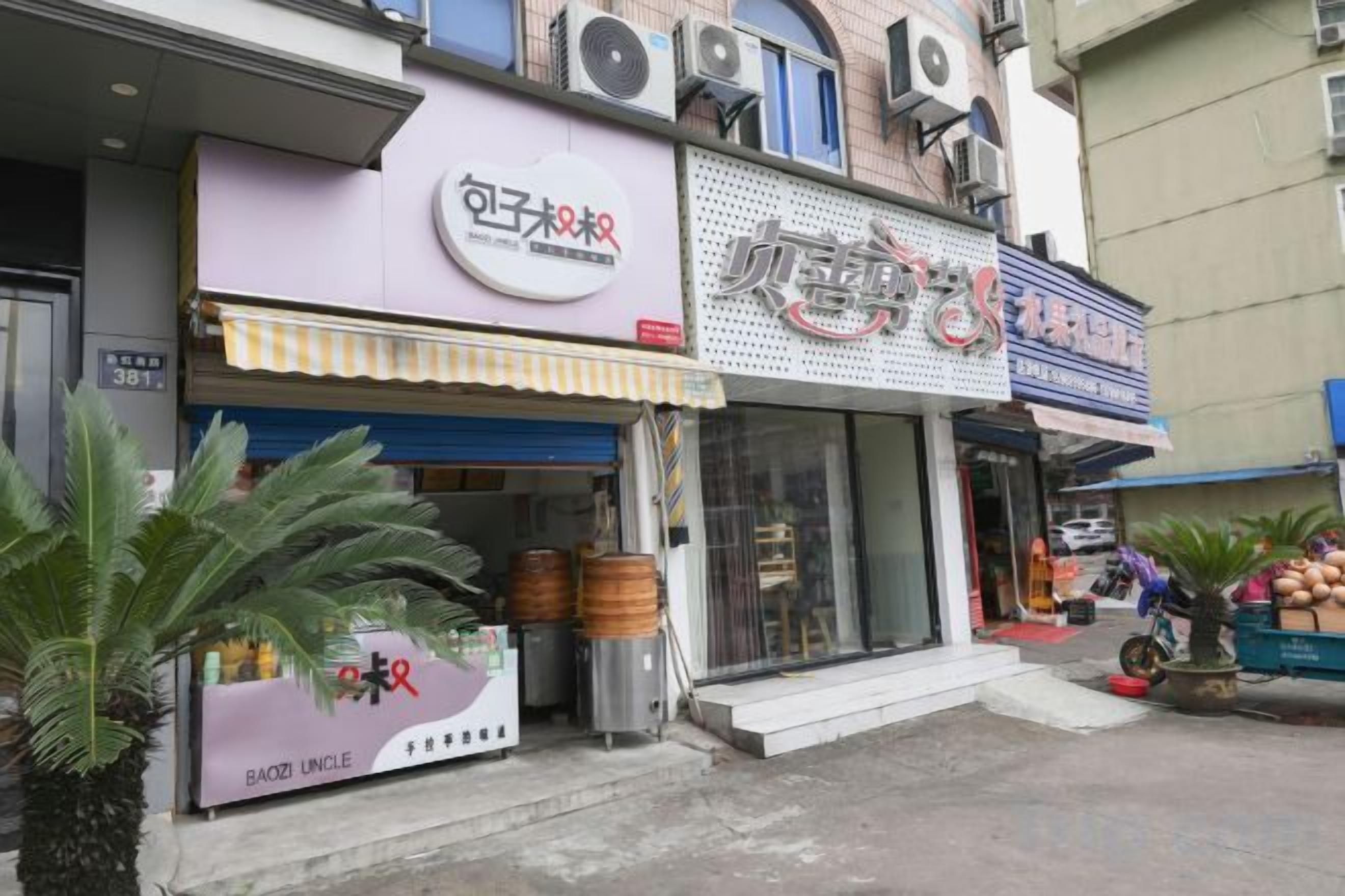Fachada com uma placa que diz 'Hanji Supermarket' e outra para 'Baodji Uncle'. Há uma exposição de produtos do lado de fora, incluindo o que parece ser uma banca de frutas.