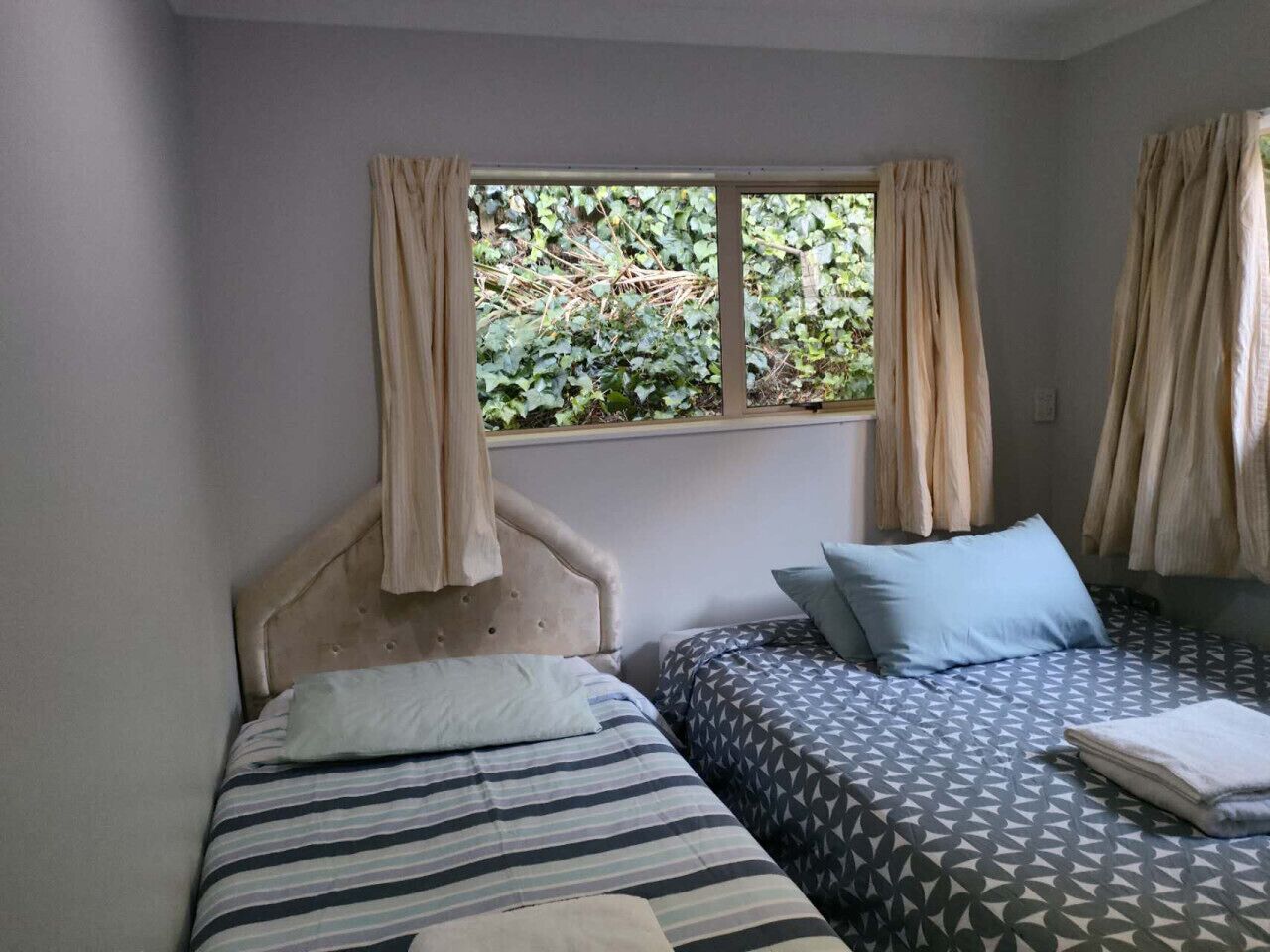 Quarto com duas camas, cortinas bege, janela com vista para vegetação e cabeceira em uma das camas.