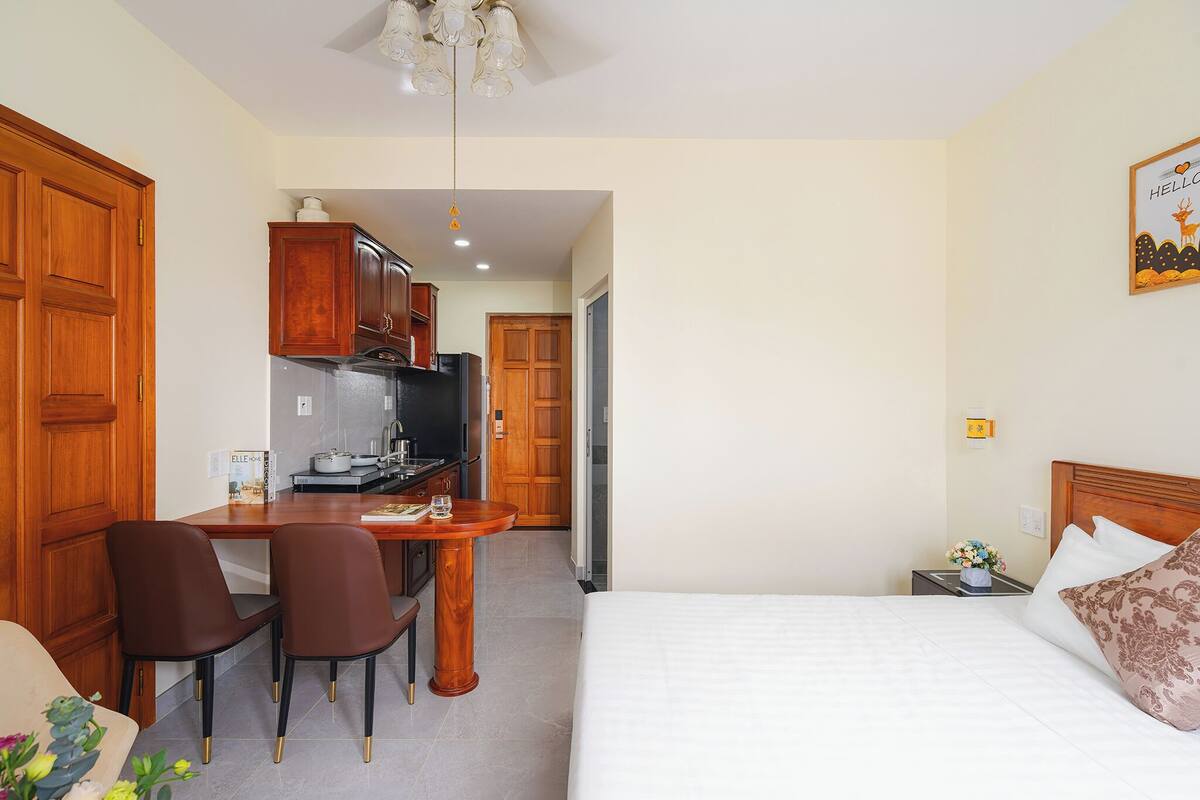 Quarto de hotel pequeno, com cama, secretária com cadeiras, cozinha compacta e guarda-roupa de madeira.