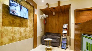 Um hall com parede de madeira, sofá cinza, mesa redonda pequena, televisão montada e balcão de recepção com folhetos.