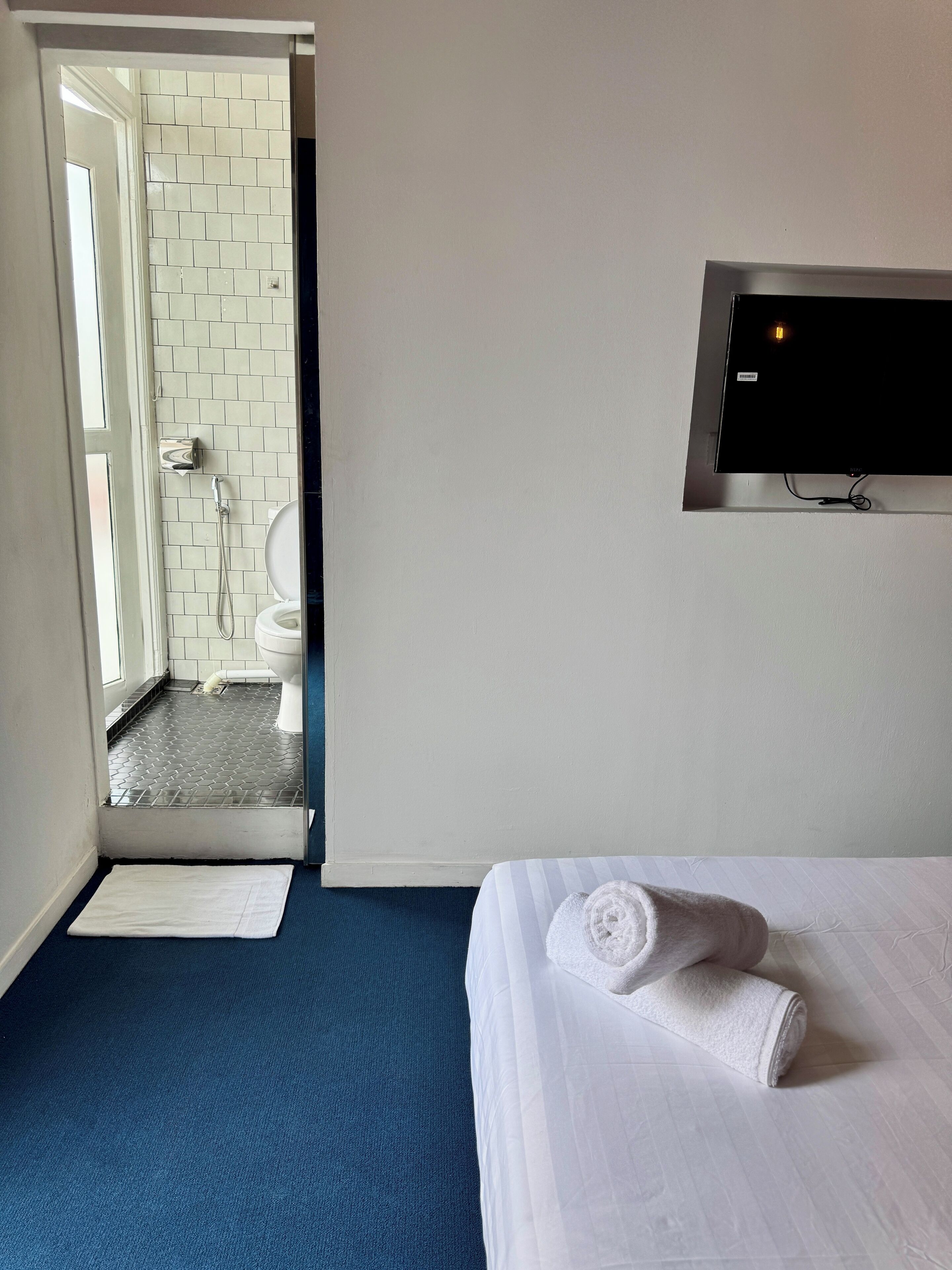 Quarto de hotel com cama, televisão presa à parede, banheiro visível por uma porta aberta e tapete azul.