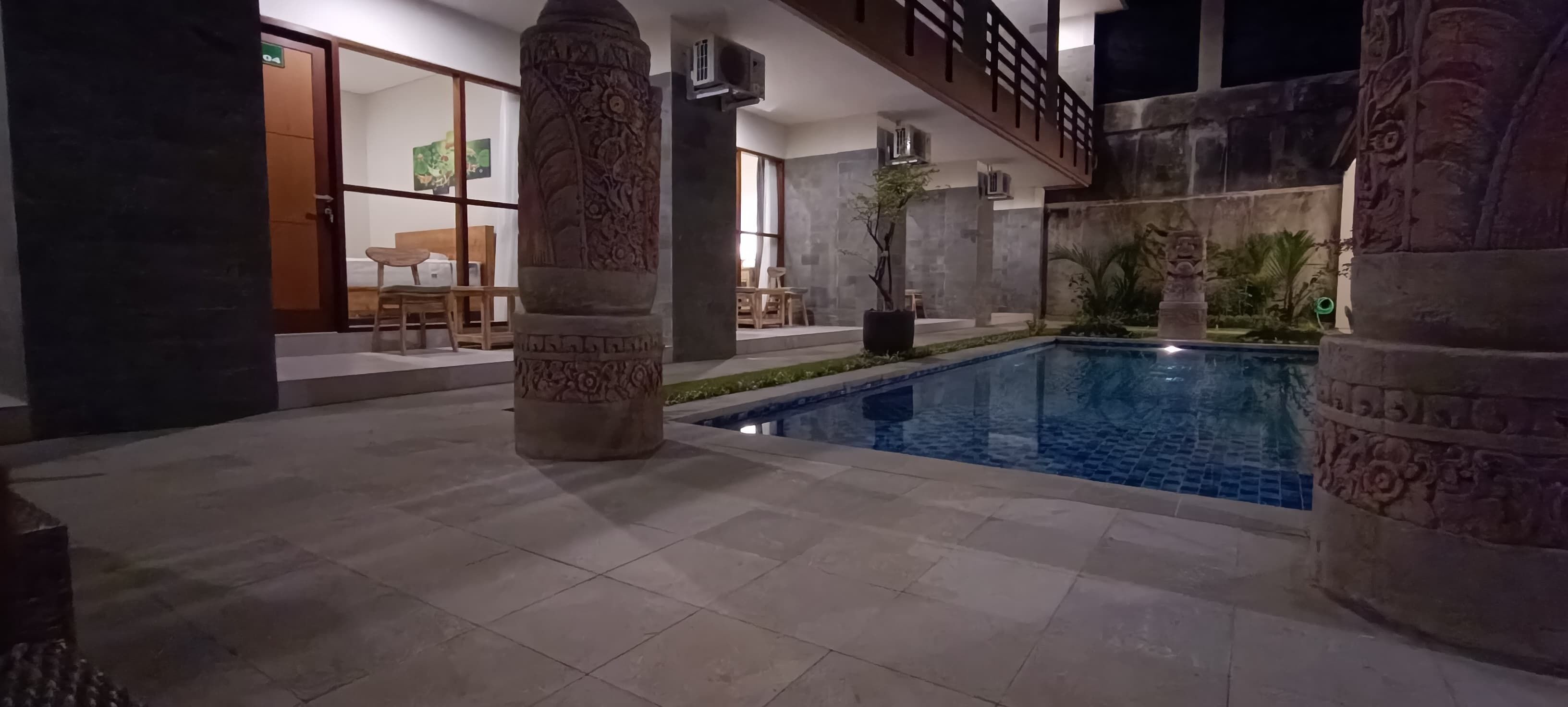Área de piscina com pilares de pedra, um deck de madeira e um espaço de estar com mesas e cadeiras.