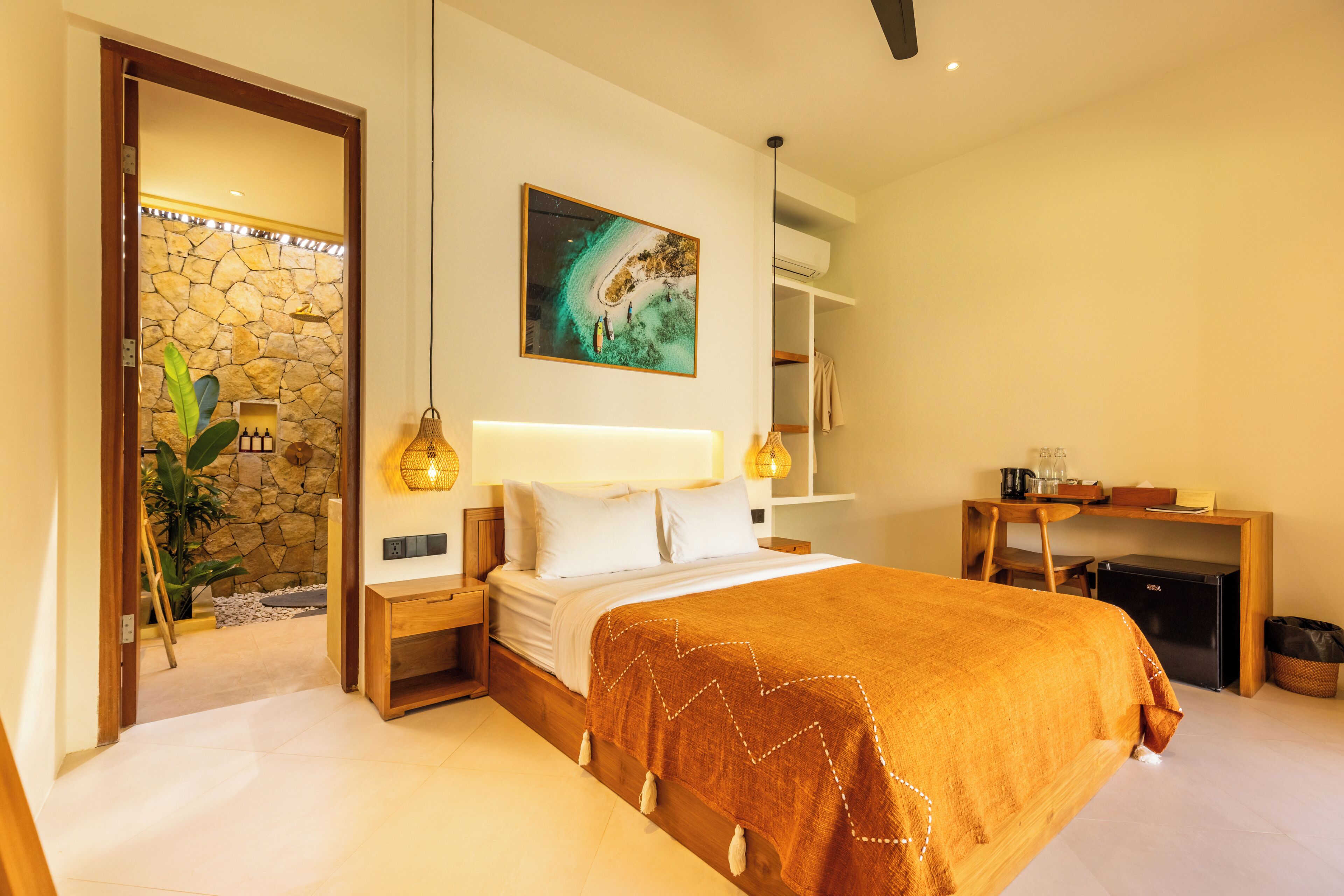 Um quarto com cama, escrivaninha, cadeira, um pequeno frigorífico e uma parede de pedra.