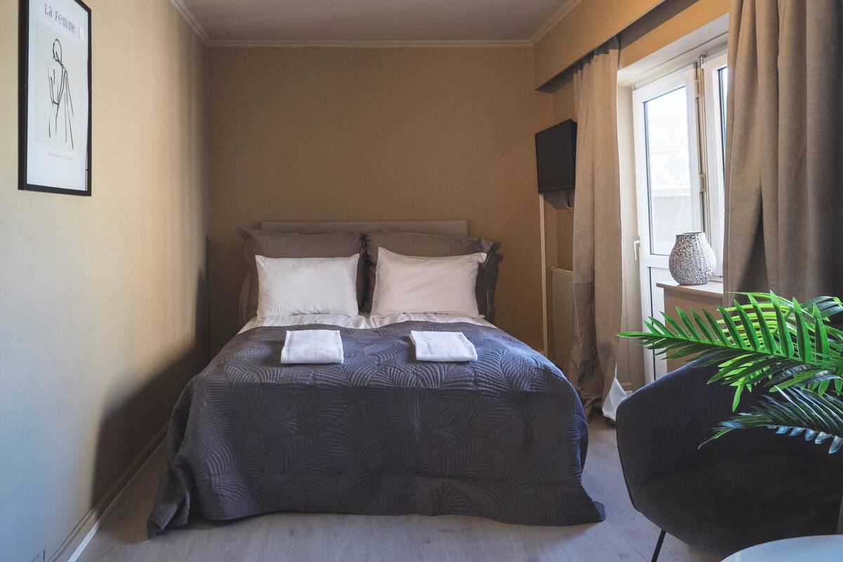 Um quarto com uma cama, uma cadeira, uma mesa pequena, uma janela com cortinas e uma televisão fixada na parede.