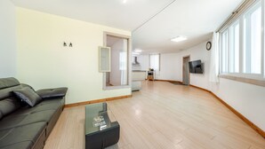 Uma sala de estar ampla com um sofá, uma mesa de centro de vidro e uma área de cozinha ao fundo.