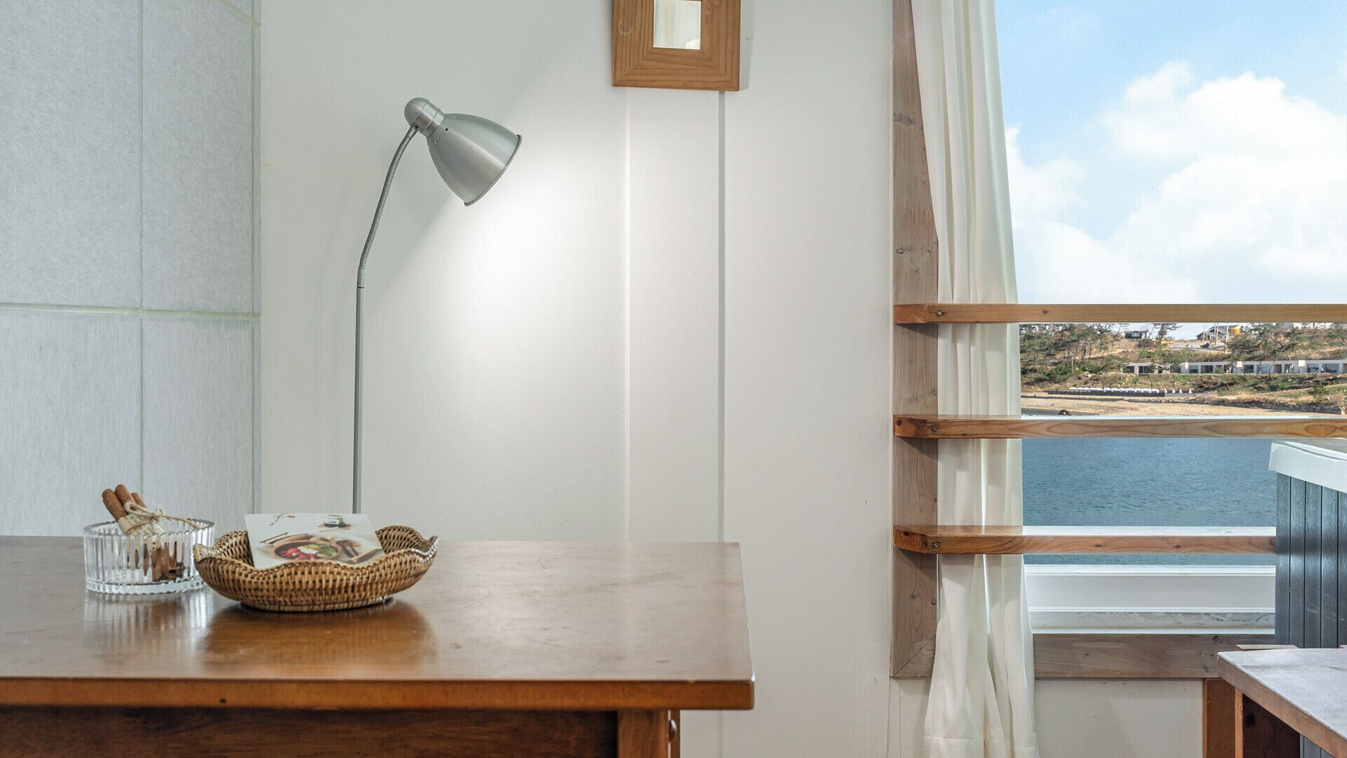 Um quarto com uma mesa de madeira, um abajur, uma cesta de vime e vista para um corpo d'água através de uma janela com molduras de madeira.
