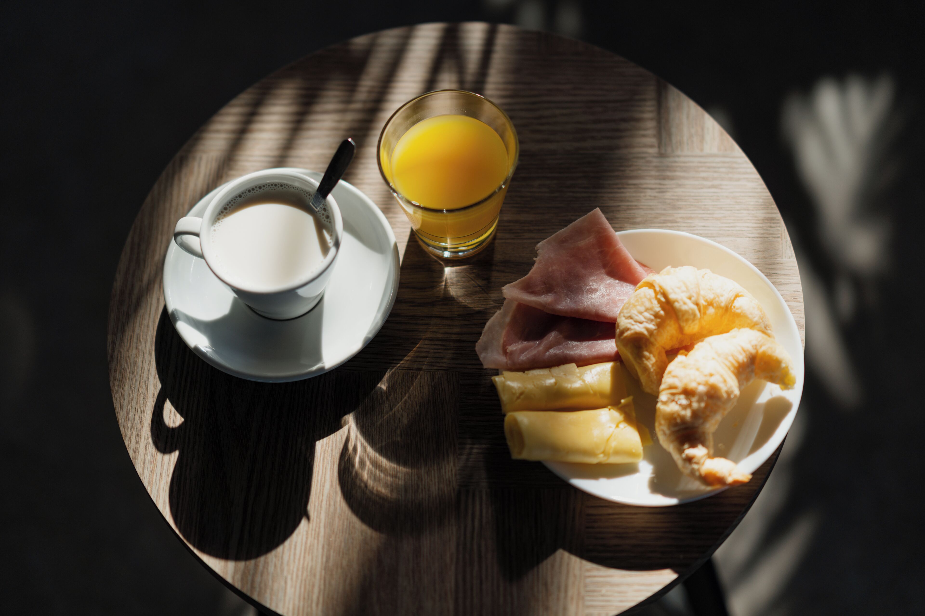 Uma mesa de madeira redonda com uma chávena e pires brancos, um copo de sumo de laranja e um prato com croissants e fiambre.