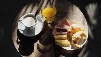 Uma mesa de madeira redonda com uma chávena e pires brancos, um copo de sumo de laranja e um prato com croissants e fiambre.