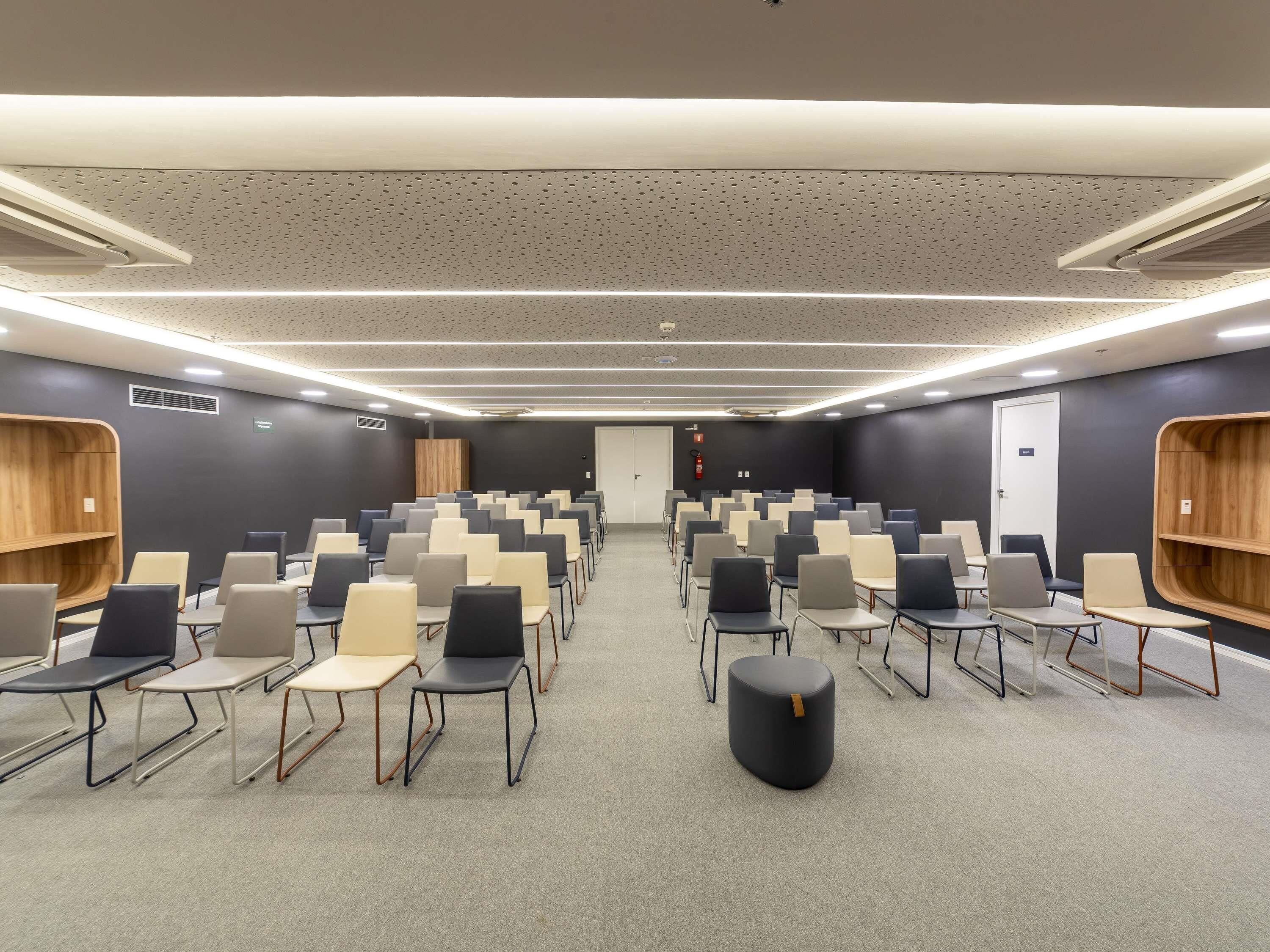 Uma sala de conferência com fileiras de cadeiras, um banquinho preto e áreas de estar em madeira.
