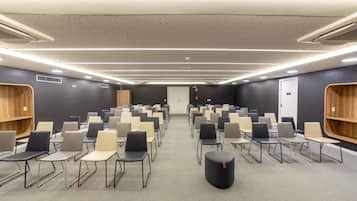 Uma sala de conferência com fileiras de cadeiras, um banquinho preto e áreas de estar em madeira.