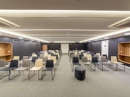 Uma sala de conferência com fileiras de cadeiras, um banquinho preto e áreas de estar em madeira.