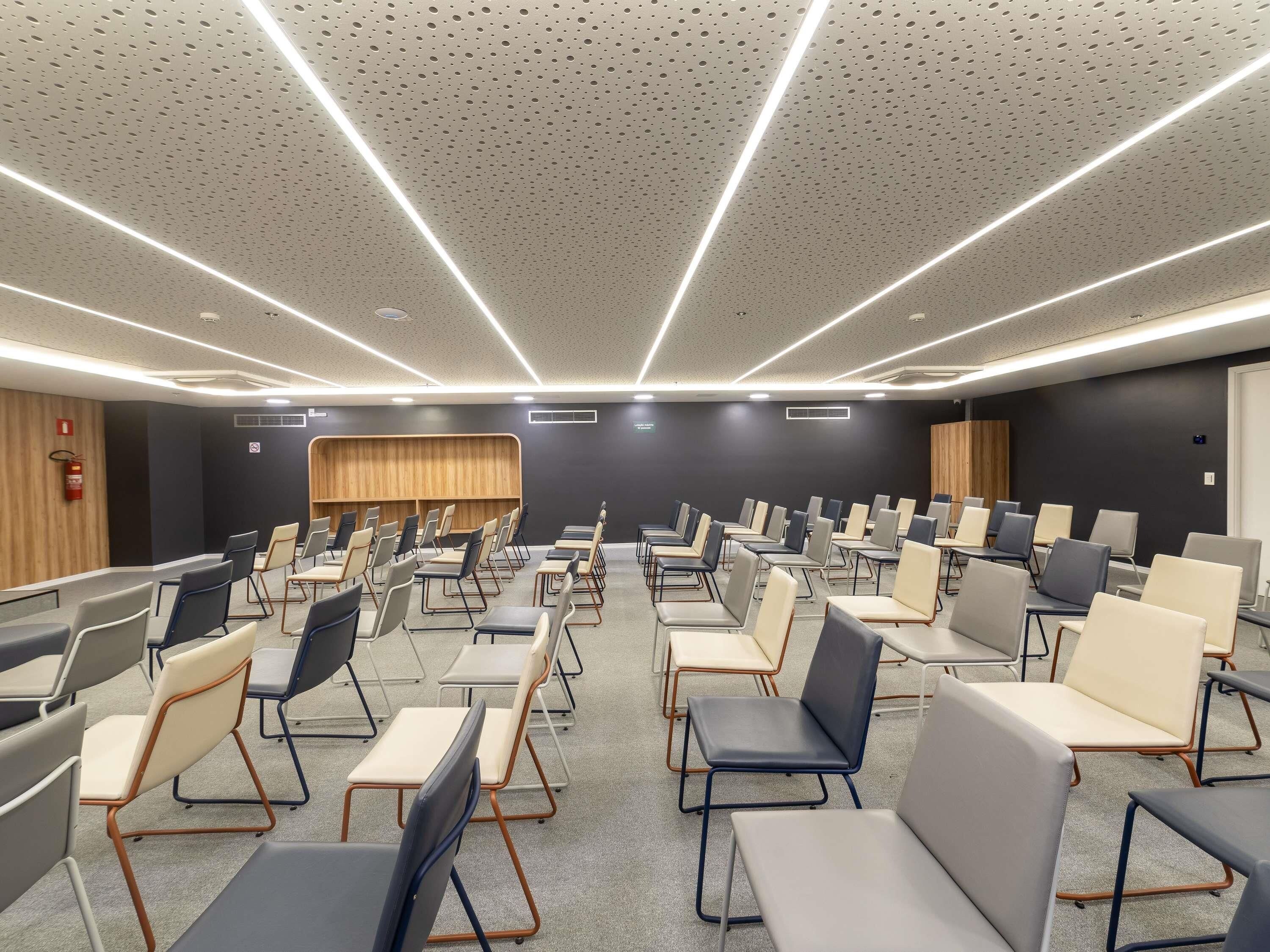 Uma sala de conferências com fileiras de cadeiras e mesas, parede de painéis de madeira e teto com design perfurado e iluminação linear.