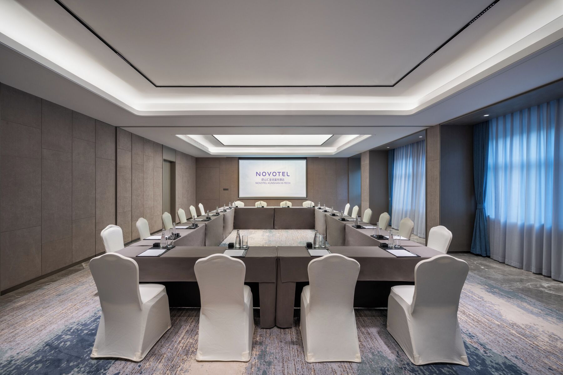 Uma sala de conferências com uma mesa comprida, cadeiras brancas e uma tela de projetor exibindo 'NOVOTEL'.