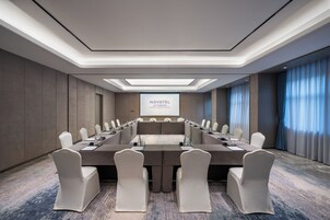Uma sala de conferências com uma mesa comprida, cadeiras brancas e uma tela de projetor exibindo 'NOVOTEL'.