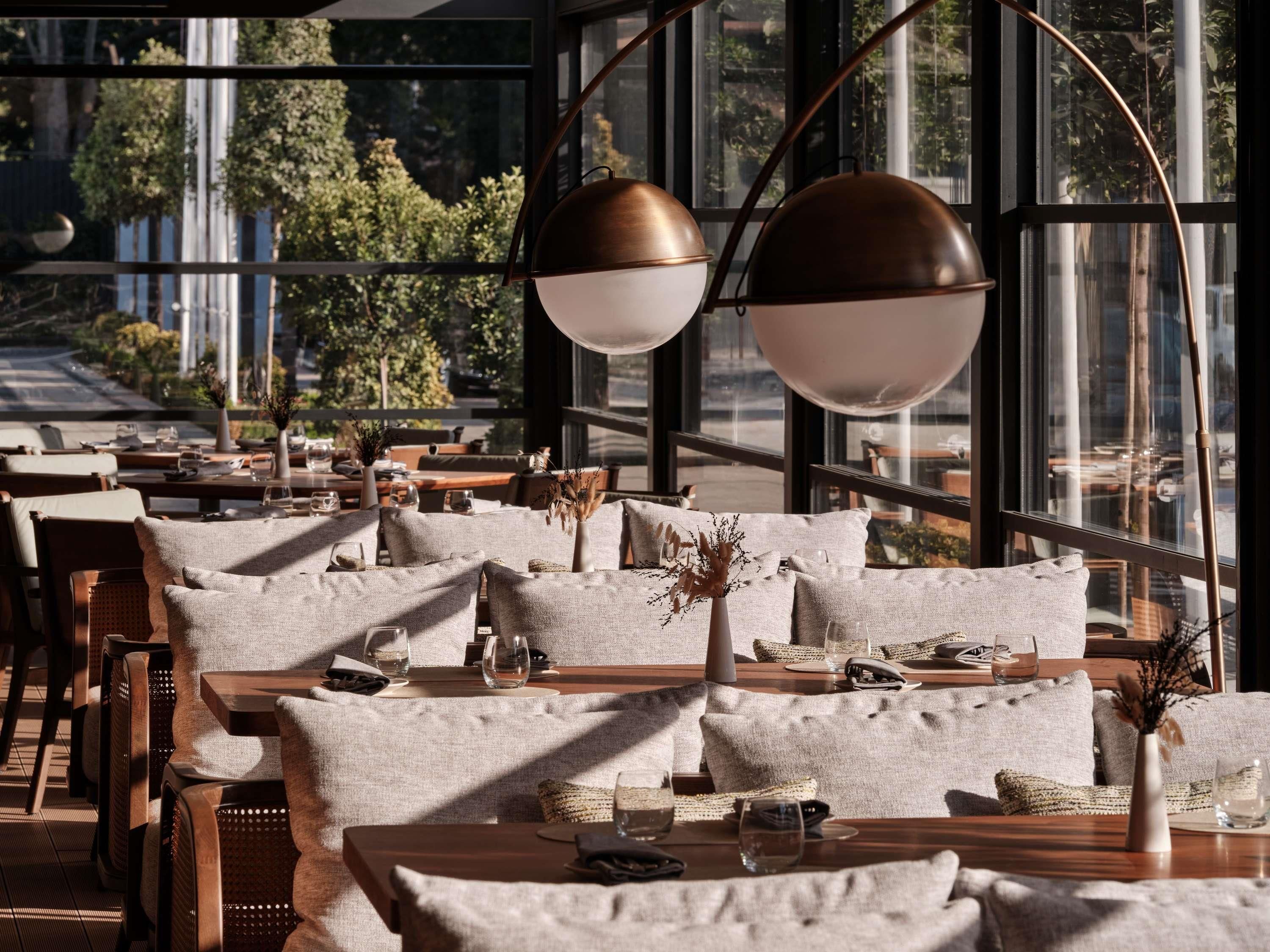 Restaurante com interior moderno, com grandes janelas, mesas de madeira e assentos confortáveis.
