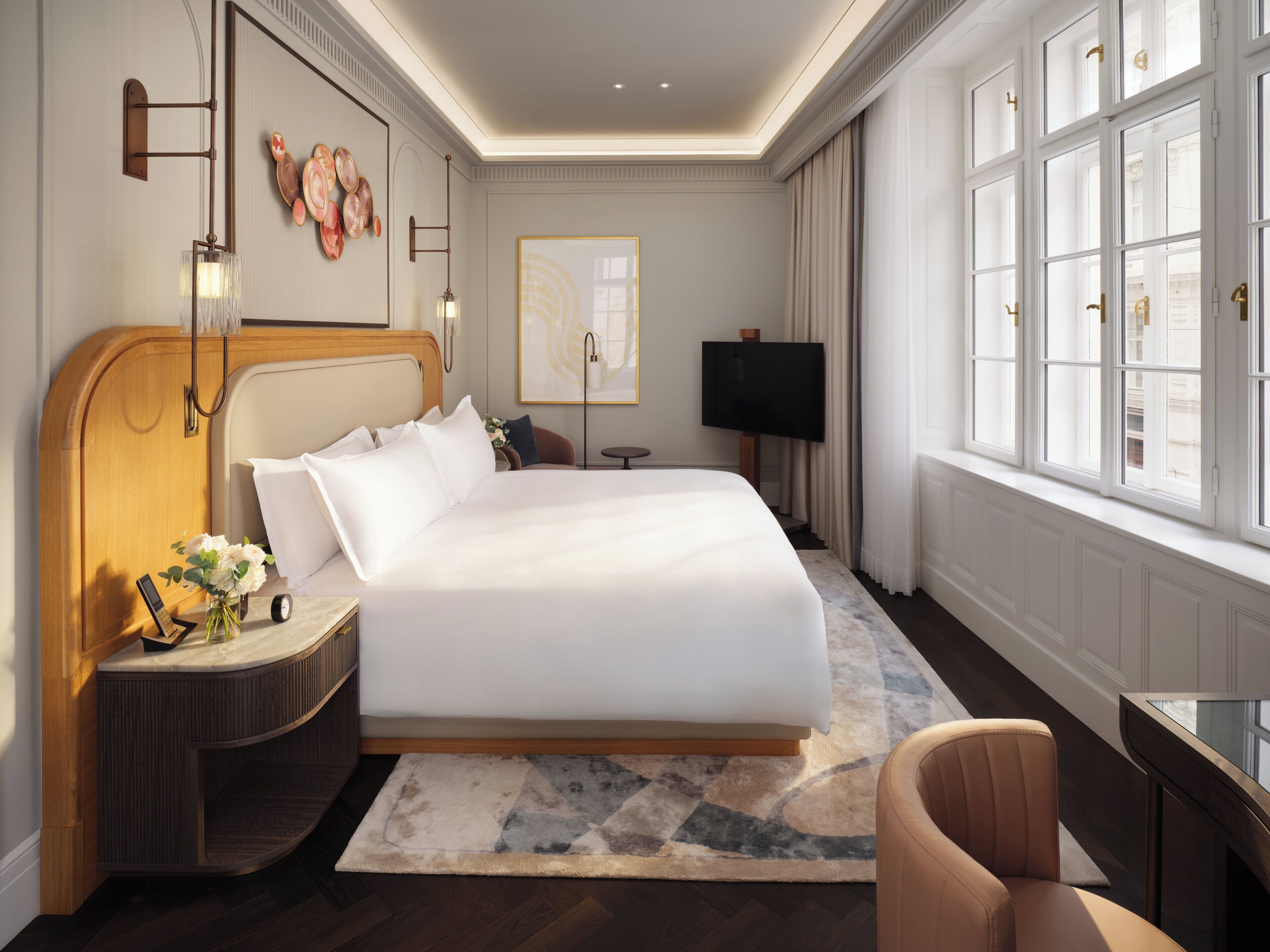Quarto de hotel espaçoso com uma cama grande, mesa de cabeceira com abajur, televisão e vista da cidade por grandes janelas.
