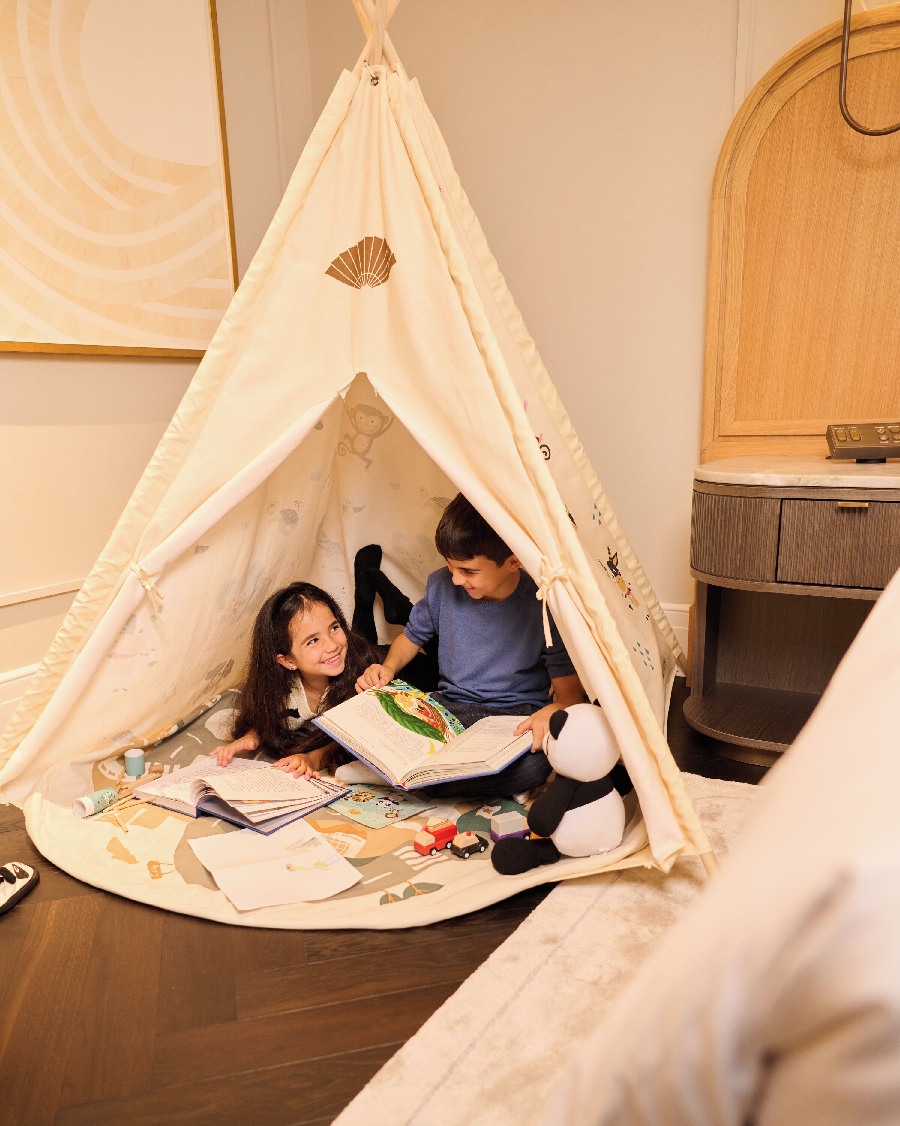 Quarto com uma cama redonda, um armário de madeira e uma pequena estrutura tipo tenda onde duas crianças leem um livro.