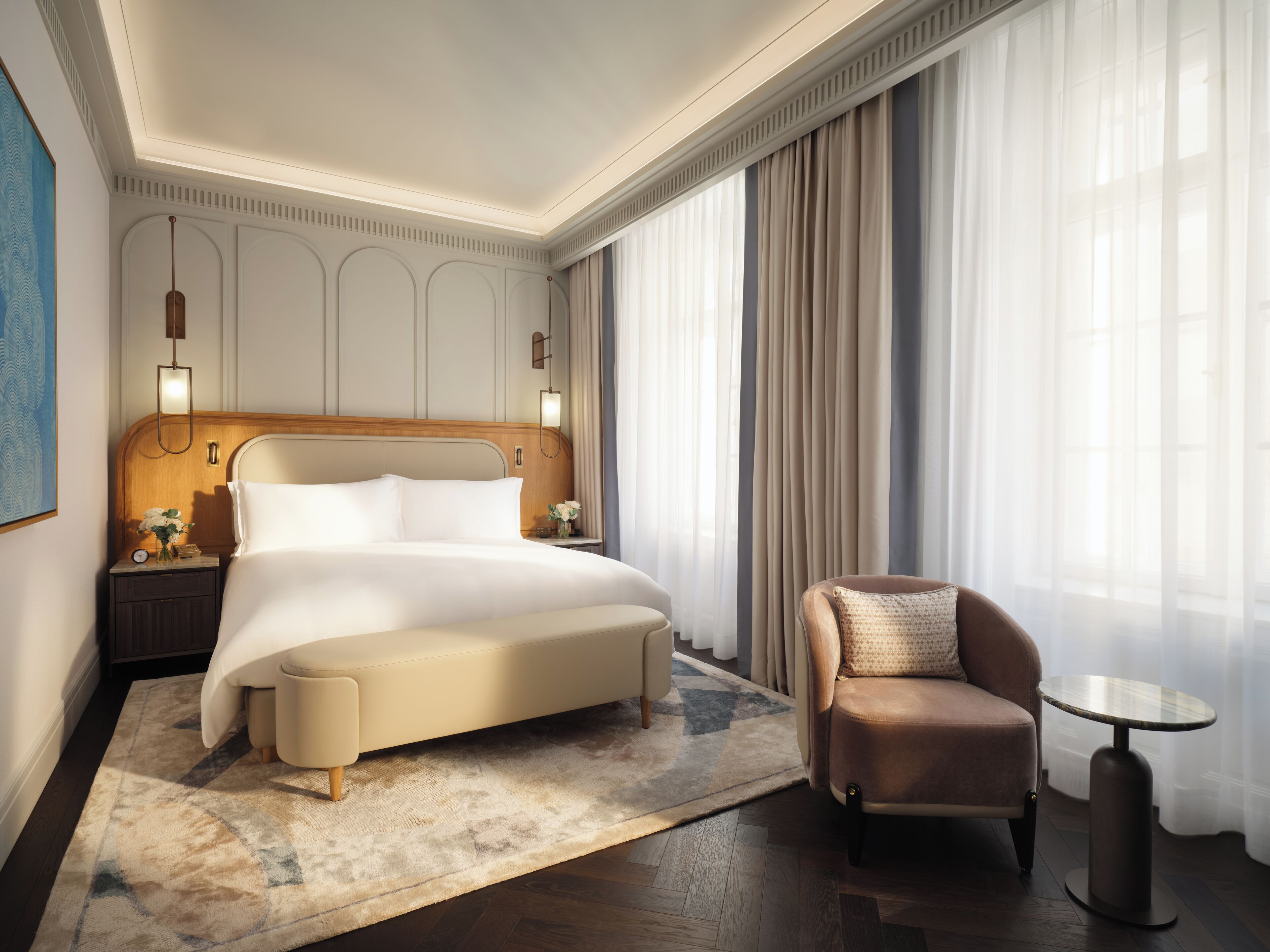 Quarto de hotel com uma cama grande, uma cadeira, uma mesinha e uma mesa de cabeceira com abajur.