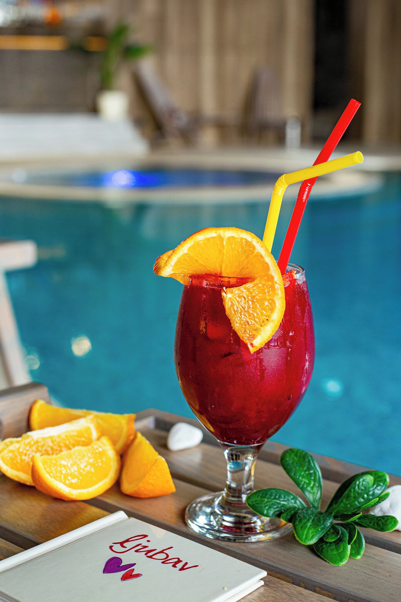 Um copo de bebida vermelha com fatias de laranja e canudos, colocado sobre uma mesa de madeira com uma piscina ao fundo.