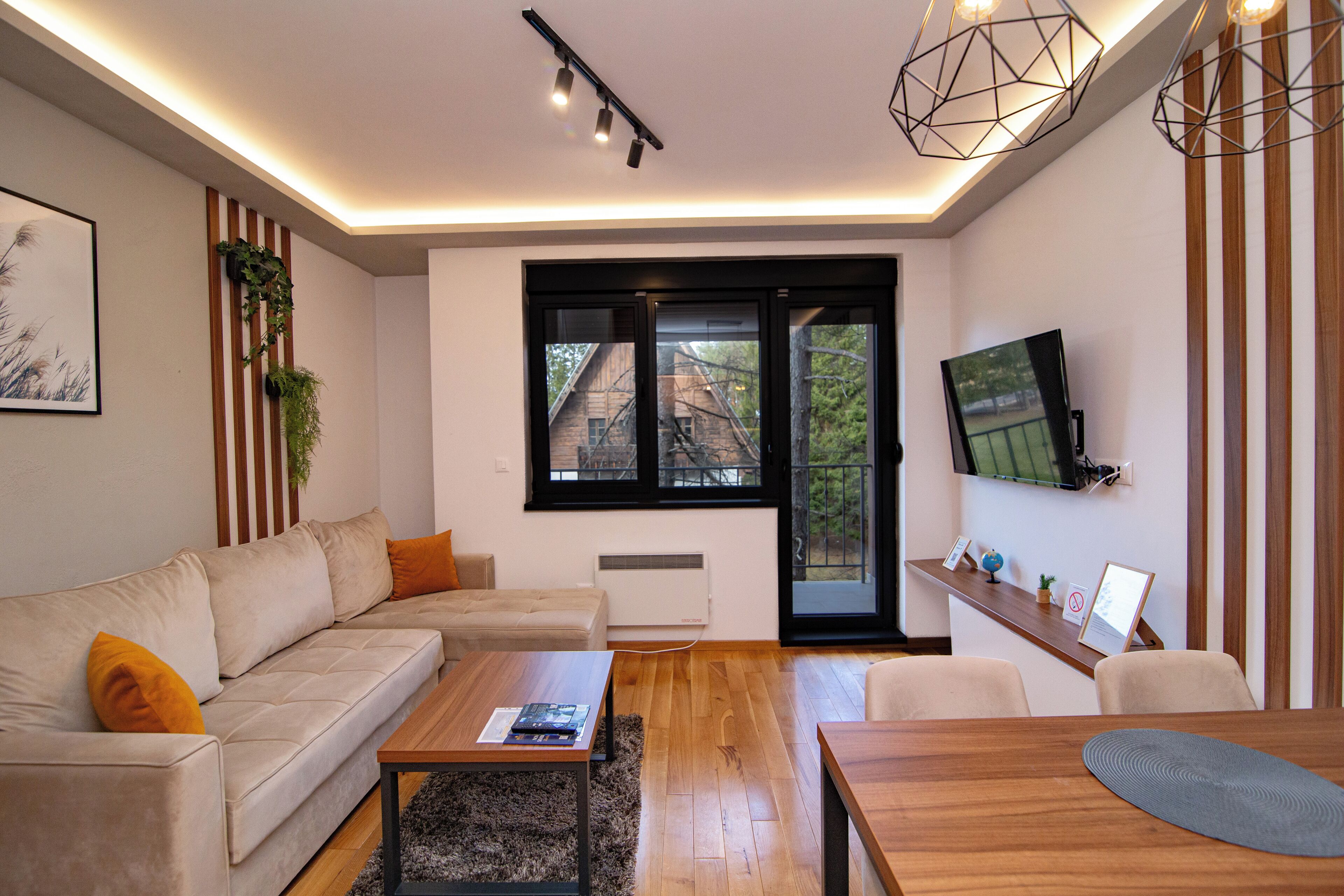Uma sala de estar moderna com sofá, mesa de centro e mesa de jantar. Há uma TV na parede, uma janela com vista para o exterior e uma porta que dá para outro cômodo.