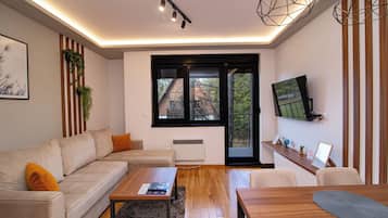 Uma sala de estar moderna com sofá, mesa de centro e mesa de jantar. Há uma TV na parede, uma janela com vista para o exterior e uma porta que dá para outro cômodo.