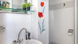 Casa de banho com lavatório branco, espelho e chuveiro. Há flores vermelhas na parede e uma prateleira de vidro com artigos de higiene pessoal.