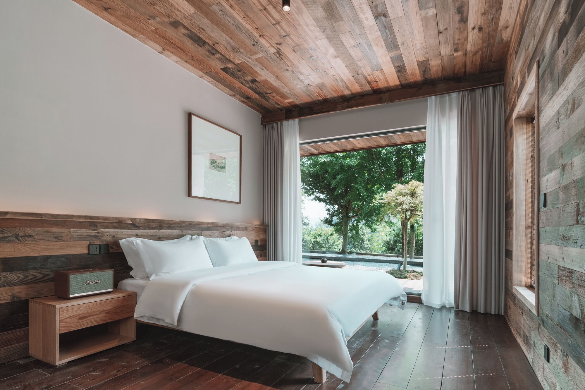 Quarto com teto de madeira, cama com roupa de cama branca, mesa de cabeceira com rádio e janela com cortinas.