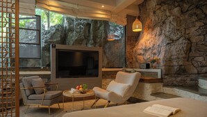 Quarto moderno com uma cama grande, uma TV, uma parede de pedra e vista para uma paisagem rochosa.