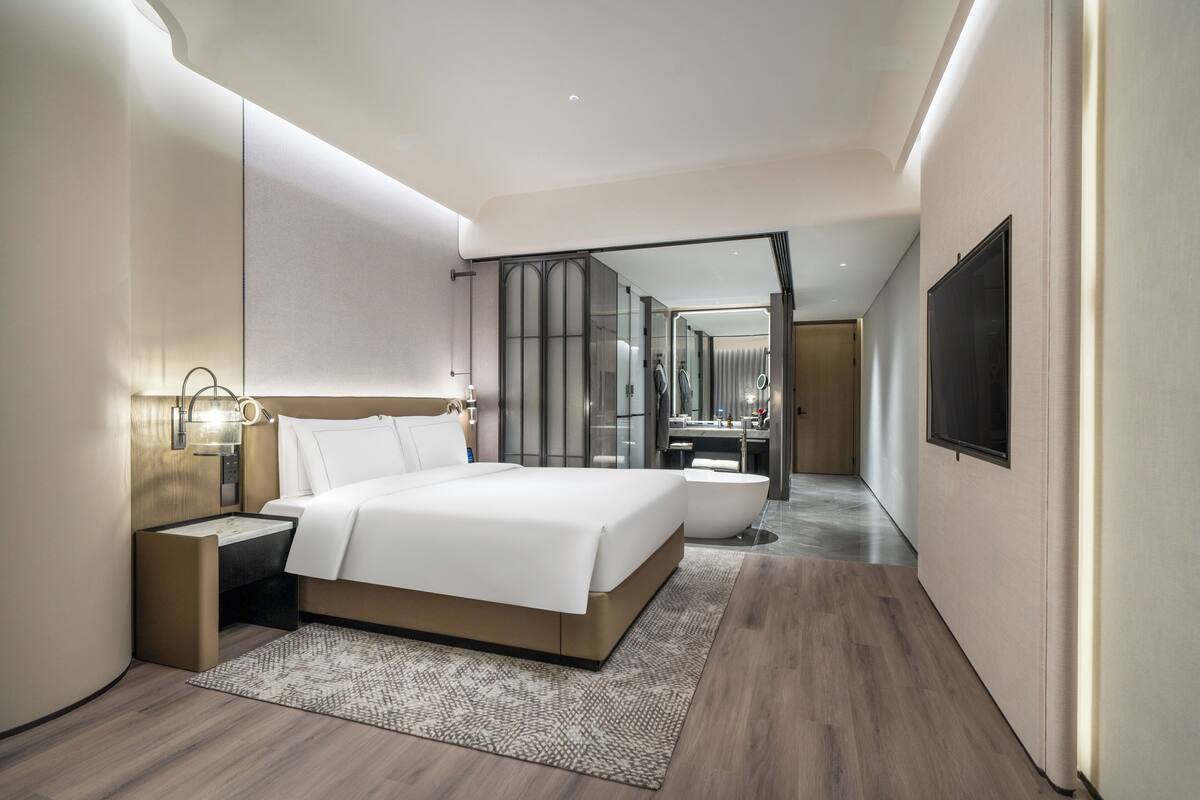 Quarto de hotel moderno com uma cama grande, uma mesa de cabeceira, televisão de tela plana na parede e um banheiro visível através de um painel de vidro.