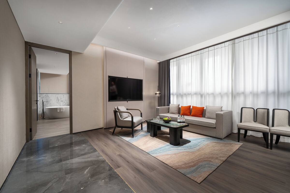 Uma sala de estar moderna com televisão de tela plana, um sofá com almofadas laranja, uma mesa de centro e uma grande janela com cortinas.