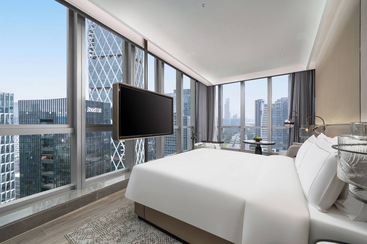 Quarto de hotel moderno com uma cama grande, uma TV de tela plana, uma pequena mesa com abajur e vista da cidade através de janelas amplas.