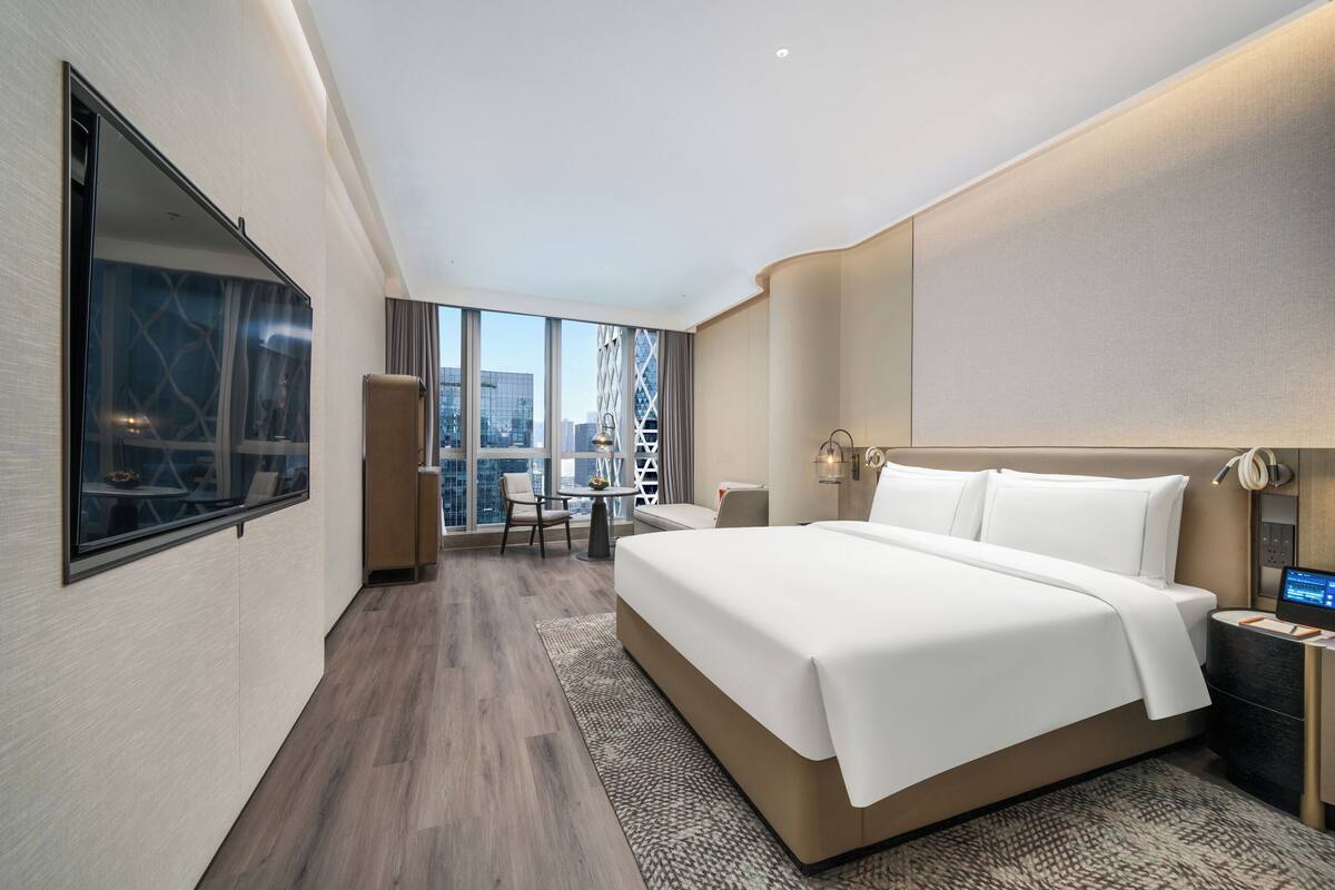 Quarto de hotel moderno com uma cama grande, uma escrivaninha com computador, uma cadeira e vista para a cidade através de amplas janelas.