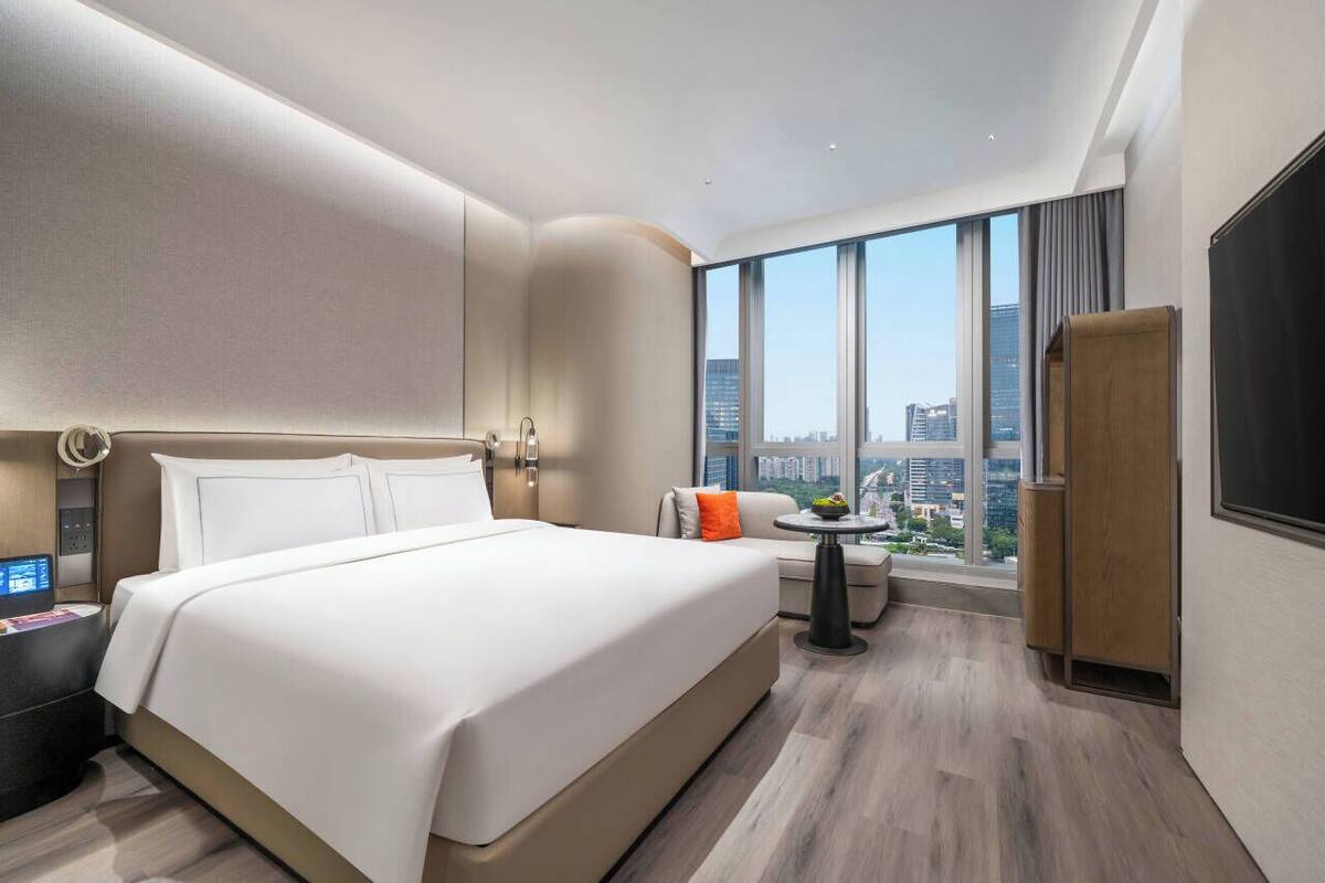 Quarto de hotel moderno com uma cama grande, uma mesa pequena, um sofá e uma televisão. O quarto oferece vista para a cidade através de grandes janelas.