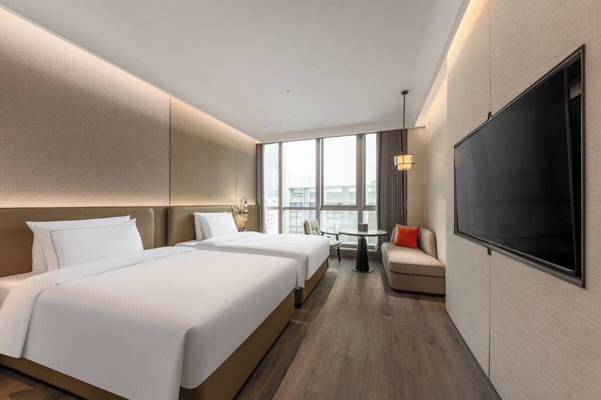 Quarto de hotel com duas camas, um sofá, uma televisão e janela com vista para a cidade.