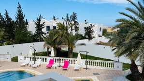 Uma área de piscina com espreguiçadeiras brancas e cor-de-rosa, céu azul límpido e edifícios brancos ao fundo.