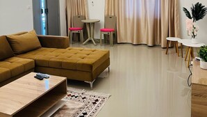 Uma sala de estar moderna com um sofá de canto, uma mesa de centro, uma pequena mesa redonda com cadeiras, um lustre e uma grande janela com cortinas.
