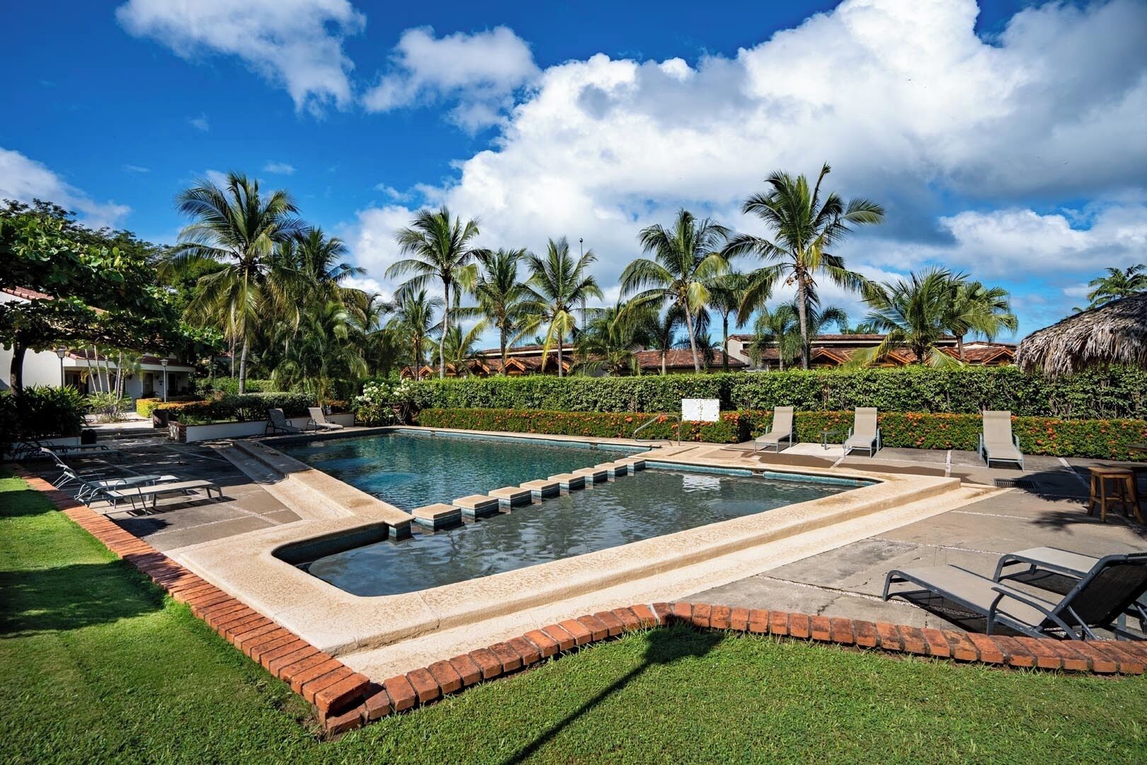 Piscina retangular com uma pequena banheira de hidromassagem, rodeada por palmeiras e espreguiçadeiras.