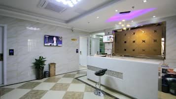 Uma área de recepção de hotel com balcão de check-in, uma televisão na parede e piso em azulejos.