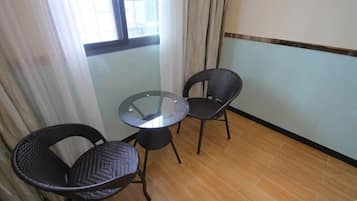 Um quarto pequeno com uma mesa de vidro e duas cadeiras, uma janela com cortinas e piso de azulejos.