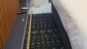Quarto de hotel moderno com uma cama, televisão, planta em vaso e uma janela grande com cortinas.