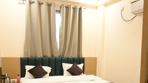 Uma cama bem arrumada com cabeceira de madeira, uma mesa de cabeceira com abajur e uma garrafa, e uma janela com cortinas.