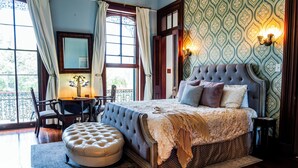 Quarto com cabeceira estofada, cama grande com várias almofadas, mesa pequena, cadeira e abajur.