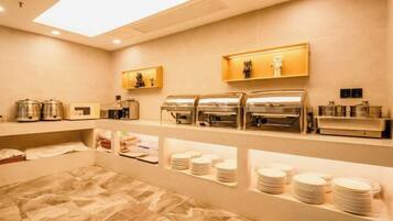 Área de buffet com distribuidores de comida, pratos e chão ladrilhado.