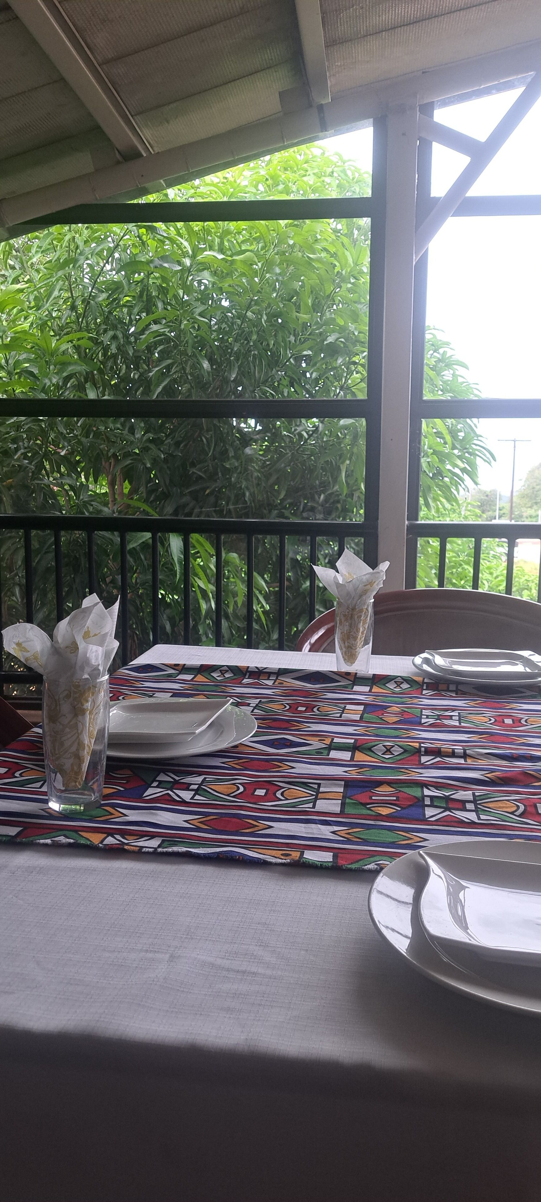 Mesa posta com pratos, copos e toalha de mesa estampada, com vista para o verde lá fora.