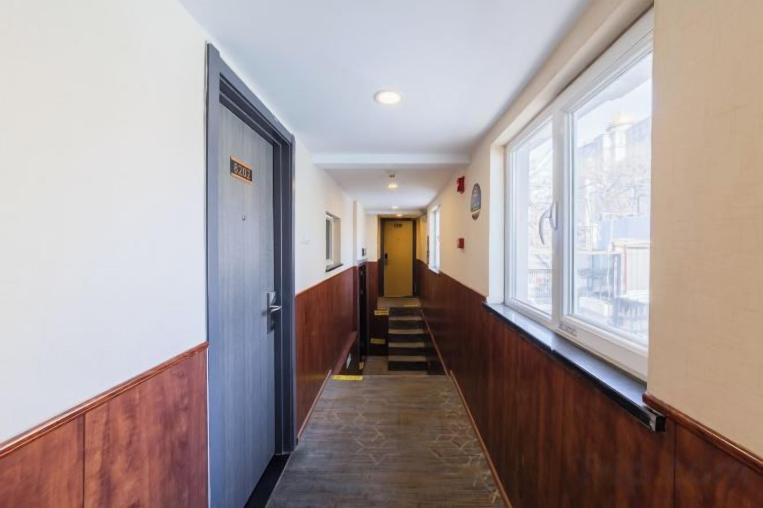 Um corredor de hotel com piso de madeira, uma porta à esquerda e janelas à direita.