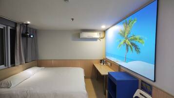 Quarto de hotel com cama, escrivaninha, televisão grande exibindo uma cena de praia, aparelho de ar condicionado e janela com cortinas.