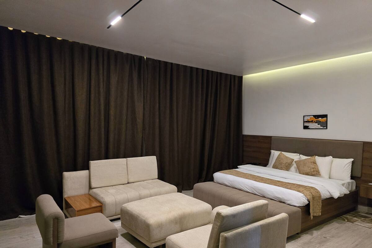 Quarto de hotel moderno com uma cama grande, duas poltronas, uma mesa pequena e uma janela ampla com cortinas.