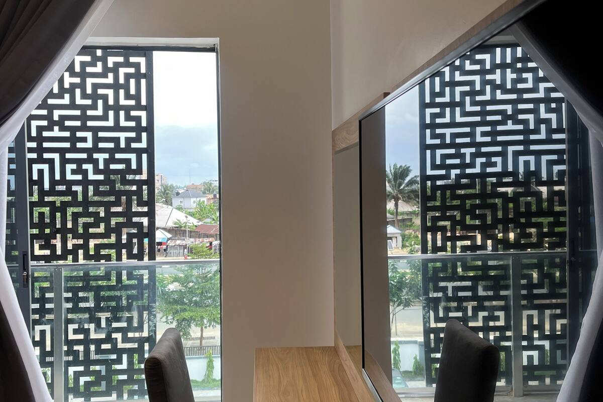 Quarto moderno com escrivaninha de madeira, duas cadeiras e uma janela grande com um painel de padrão geométrico.